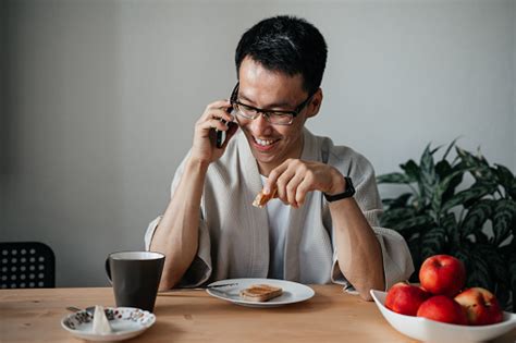 미소 짓는 성인 아시아 남자는 아침 식사 중에 스마트 폰으로 이야기하는 안경을 쓰고 있습니다 가정 생활에 대한 스톡 사진 및
