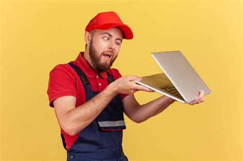 파란색 유니폼과 빨간 모자를 쓴 수염을 가진 노동자 남자가 반쯤 열린 노트북을 들고 감시하고 있습니다 유머에 대한 스톡 사진 및 기타 이미지 Istock