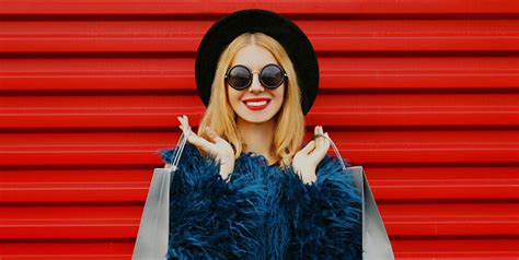 파란 인조 모피 코트를 입은 두 개의 쇼핑백과 빨간 벽 배경 위에 검은 둥근 모자를 쓰고 있는 세련된 금발 여성의 초상화 클로즈업 25 29세에 대한 스톡 사진 및 기타