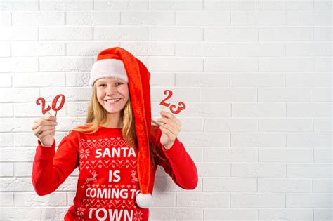 빨간 스웨터와 산타 모자를 쓴 어린 소녀가 텍스트를 위한 장소가 있는 흰색 벽돌 벽에 2023이라는 숫자를 손에 들고 있습니다 12 13세에 대한 스톡 사진 및 기타 이미지