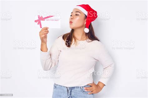 산타클로스 모자를 쓴 쾌활한 젊은 아시아 여성 20대가 흰색 스튜디오 배경에 격리된 손에 상품권 쿠폰 카드를 들고 키스합니다 메리 크리스마스 컨셉 25 29세에 대한 스톡