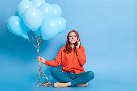 캐주얼 한 스웨터와 청바지에 행복한 젊은 빨간 머리 여자 소녀 푸른 배경에 고립 포즈 생일을 축하하는 여자 헬륨 풍선을 들고 휴대 전화로 이야기 20 24세에 대한 스톡