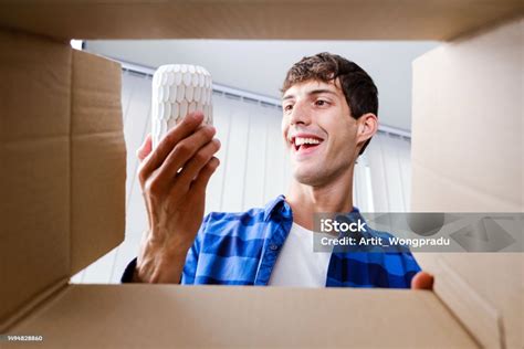 상자 안에서 바라보는 미소 짓는 남자는 소포를 풀고 꽃병을 들고 배경에 커튼을 치고 앉아 온라인 상점 주문으로 골판지 상자를 여는 행복한 고객을 만족시켰습니다 25 29세에