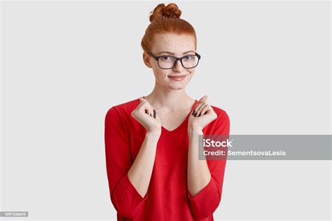 머리 매듭 기쁘게 빨간 머리 여 자가 투명 안경을 착용 기쁨을 표현 하 고 뭔가 대 한 예상 빨간색 점퍼를 입은 가슴 위에 손을 유지 화이트 스튜디오 벽에 대 한 모델