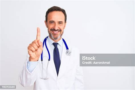 외투와 청진기를 입은 중년 의사는 자신감과 행복한 미소를 지으면서 손가락 번호 1을 보여주고 가리키는 고립 된 흰색 배경 위에 서 있습니다 가리키기에 대한 스톡 사진 및
