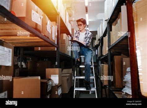 Immersed Female Warehouse Worker Sitting On The Ladder Writing On A