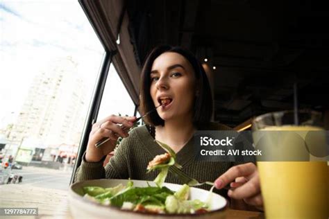 갈색 머리 여자 먹는 시저 샐러드 와 즐거움 전면에 오렌지 신선한 주스의 흐린 유리 어두운 배경과 큰 빛 창 비즈니스 점심 시간 개념 맛있고 건강한 음식 20 24세에 대한