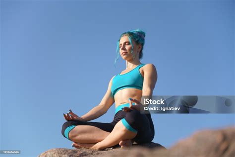 청록색 검은 요가 의상을 입은 청록색 머리를 가진 매력적인 아름다운 젊은 여성은 하늘을 향해 돌에 다리를 꼬고 똑바로 뒤로 눈을 감고 무릎에 손을 대고 상징적 이거나