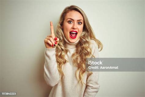 고립 된 흰색 배경 위에 겨울 터틀넥 스웨터를 입은 아름다운 여성이 성공적인 아이디어로 손가락을 가리킵니다 종료하고 행복 넘버 원 가리키기에 대한 스톡 사진 및 기타 이미지