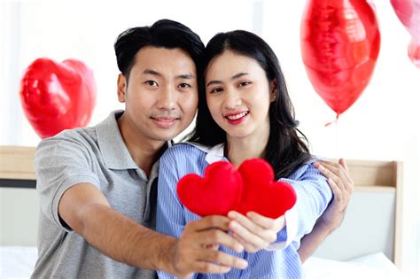 침실에서 빨간 하트를 들고 카메라에 보여주는 행복한 아시아 커플 하얀 침실에서 발렌타인 데이에 기억에 남는 기념일을 축하하는 낭만적인 연인 사랑 축하 개념 2명에 대한 스톡
