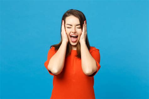 아름 다운 행복 한 젊은 여성이 빨간 오렌지 드레스를 입고 얼굴에 손을 넣어 푸른 벽 배경 스튜디오 초상화 위에 고립 승리에 대해 발견 했다 사람들의 라이프 스타일 패션