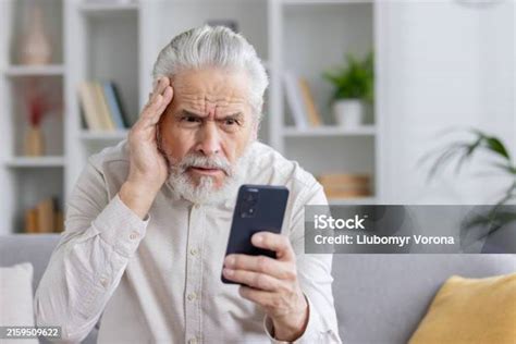 충격에 휩싸인 노인이 집에서 흰 셔츠를 입고 머리를 감싸 안은 채 걱정스러운 표정으로 휴대전화 화면을 바라보는 클로즈업 사진 Brand Name Smart Phone에 대한
