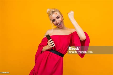 빨간 드레스를 입은 유럽 금발 소녀는 복사 공간이있는 노란색 스튜디오 배경에 휴대 전화를 들고 기쁨으로 미소지었습니다 스마트폰에 대한 스톡 사진 및 기타 이미지 Istock