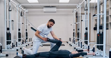 치료실에서 재활 장비의 밧줄이 부착된 무릎을 구부리는 젊은 여성 물리 치료사는 저항 밴드가 부착 된 여성 환자의 오른쪽 다리를 당깁니다 회복 클리닉 치료 사진