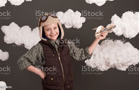 한 소녀가 골판지 비행기를 가지고 놀고 조종사가 되는 꿈을 꾸고 복고풍 스타일의 재킷과 안경을 쓰고 면모 구름 회색 배경 갈색으로 착색된 헬멧을 입고 있습니다 갈색에 대한