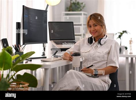 Young Female Developer Sitting Front Of Computers With Coded Data On Screen And Smiling To