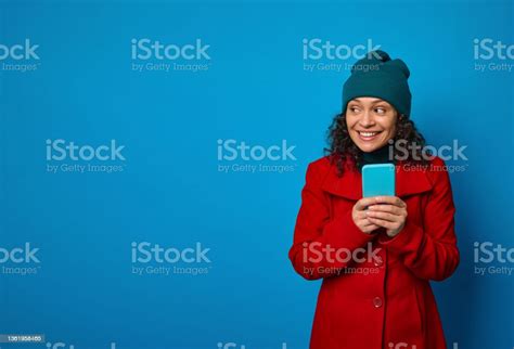 밝은 빨간 코트와 따뜻한 녹색 모직 모자를 쓰고 있는 쾌활한 행복한 예쁜 여성의 파란 배경에 고립된 초상화가 전화와 옆으로 바라보는 것을 기뻐합니다 광고 공간 복사 사람들에