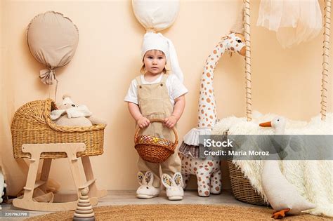 토끼 귀가 달린 모자를 쓴 쾌활한 아기가 부드러운 깔개 위에 앉아 형형색색의 부활절 달걀로 가득 찬 고리버들 바구니를 들고 있습니다 계절에 대한 스톡 사진 및 기타 이미지