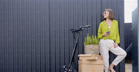 회색 벽 배경에 소포와 화분이 있는 전기 스쿠터 근처에 전화기를 들고 서 있는 젊은 여성 배달과 지속 가능한 라이프 스타일의 개념 사진 Unsplash의 재생 가능