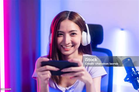 휴대 전화에서 비디오 게임 승리 거실에서 스마트 폰을 들고 의자에 앉아있는 젊은 아시아 예쁜 여자 행복한 여성 프로 스트리머 중국어 착용 헤드폰 네온 라이트와 온라인 게임을