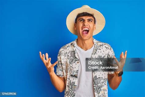 인도 남자 휴가 하와이안 셔츠 여름 모자 를 입고 고립 된 파란색 배경 미친 미친 미친 소리와 공격적인 표정과 팔을 제기 고함 좌절 개념입니다 감정에 대한 스톡 사진 및