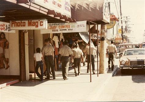 Several People Are Walking Down The Sidewalk In Front Of A Store With Cars Parked On The Street