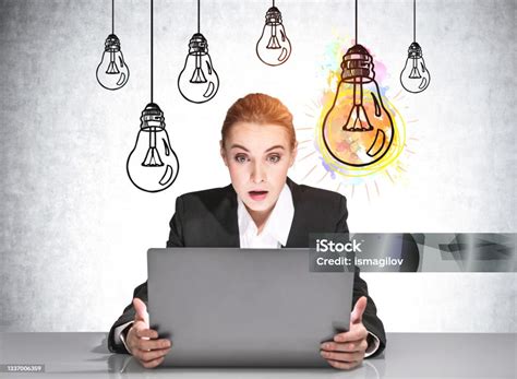 노트북 앞 책상에 앉아 경력 기회에 대해 궁금해 하는 양복을 입은 매력적인 비즈니스 여성의 초상화 뒤에 벽에 그려진 노란색 전구 개념에 대한 스톡 사진 및 기타 이미지