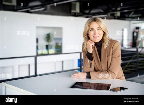Blond Female Professional With Hand On Chin Leaning On Desk In Office