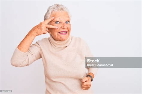 고립된 흰색 배경 위에 서있는 터틀넥 스웨터를 입은 선임 회색 머리 의 여성 얼굴에 손가락으로 평화 기호를하고 승리를 보여주는