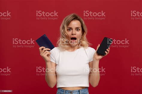 빨간 배경에 흰색 티셔츠에 곱슬 금발 머리를 가진 여자의 초상화입니다 깜짝 놀란 모델은 카메라를 보고 휴대 전화로 여권을 들고