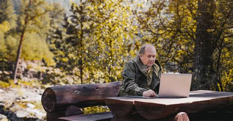 작업복을 입은 성숙한 남자가 공원의 나무 테이블에 앉아 노트북 작업을 하고 있습니다 헌터 포레스터는 벤치에 앉아 흐릿한 배경에 언덕과 나무가 있는 넷북을 사용하고 있습니다