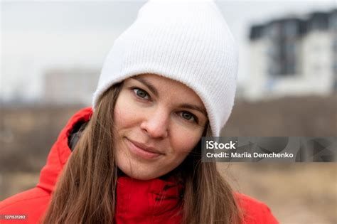 하얀 니트 모자와 흐린 배경에 대한 흐린 날에 거리에서 빨간 재킷을 입고 긴 머리를 가진 아름다운 젊은 여성을 미소 짓는 메이크업 자연의 아름다움이없는 여성 클로즈업 보기