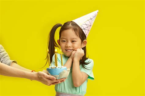 휴일의 분위기 파티 종이 모자를 쓴 사려 깊은 아이 소녀는 케이크에 생일 촛불을 바라 보며 소원을 빌 준비가되어 있으며 햇볕이 잘 드는 노란색 배경에 기대를 걸고 그들을