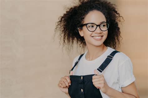 곱슬 머리를 가진 기뻐하는 아프로 미국 십대의 사진 광범위하게 미소 멀리 쳐다 보며 투명한 안경과 바지를 입고 좋은 분위기를 가지고 친구들과 자유 시간을 보내고 있습니다