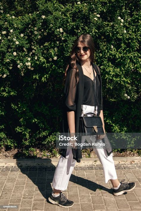 화창한 날에 꽃 덤 불에 걷는 아름 다운 머리를 가진 아름 다운 세련 된 Hipster 소녀 정원에서 하루를 즐기고 유행 복장에 선글라스와 화려한 Boho 여자 가방에 대한