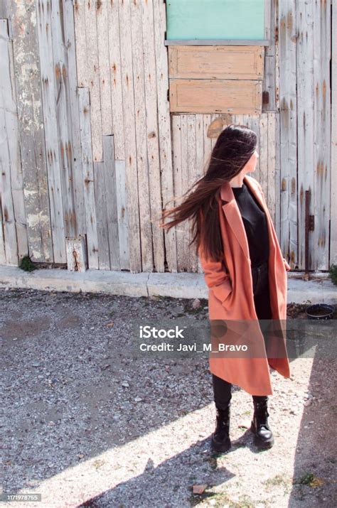 트렌치코트 주머니에 손을 얹은 젊은 여성이 그림자 사이와 나무 벽면 앞에서 빛의 공간에 서 있다 가리키기에 대한 스톡 사진 및 기타 이미지 Istock