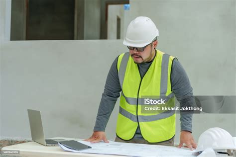 아시아 건축가 청사진과 노트북에서 건설 계획을 확인합니다 근로자를 지시하는 계획 녹색 반사 조끼와 안전모를 쓴 남자들 건설 현장의 업무용 책상에서 35 39세에 대한 스톡