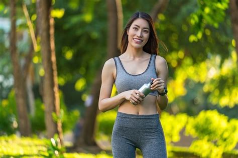 스마트워치를 들고 스포츠복을 입은 젊은 아시아 여성이 스포츠 물병을 들고 공원에서 여유롭게 산책을 즐기며 따뜻한 일몰 빛 속에서 건강한 몸매를 보여줍니다 20 29세에 대한