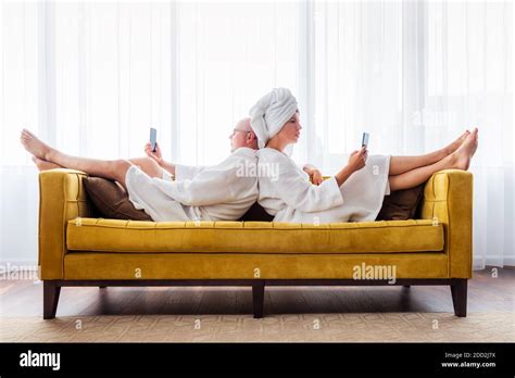 Couple In Bathrobes Is Relaxing On The Yellow Sofa With Mobile Phones