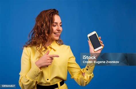 긴 곱슬 머리빈 화면 휴대 전화를 들고 파란색 배경 위에 손가락을 가리키는 행복한 아름다운 젊은 여성 20 24세에 대한 스톡 사진 및 기타 이미지 Istock