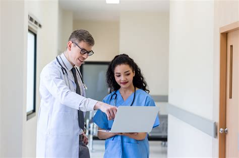 균일 한 서있는 임상의와 컴퓨터 모니터를보고 의료 문서 및 컨설팅에 대해 논의합니다 의료 사무실에서 노트북으로 작업하는 의사와