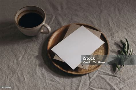 빈 인사말 카드 초대장 모형 나무 접시에 담긴 봉투의 머리 위 보기 커피 한 잔 올리브 가지 햇빛에 노출되는 린넨 테이블 천 변덕스러운 여름 아침 식사 정물 문구 작업 공간