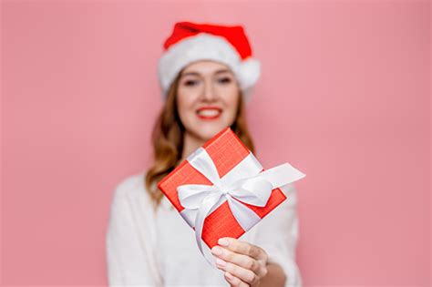 크리스마스 축제 모자 쇼에서 행복한 흐릿한 젊은 여성이 분홍색 스튜디오 배경에 고립 된 흰색 리본이있는 빨간 선물 상자를 카메라로 보여줍니다 30 34세에 대한 스톡 사진