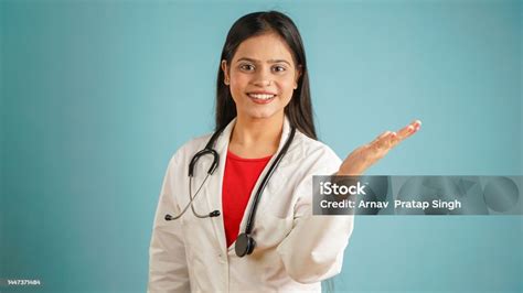 복사 공간을 손가락으로 가리키는 젊은 여성 의사의 초상화 앞치마를 입은 쾌활한 아시아 인도 여성 의사 파란색 스튜디오 배경 위에 격리된 빈 복사 공간을 보여주는 청진기