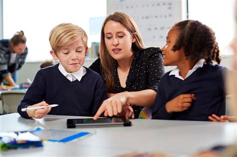 여성 학교 교사 초등 학교 교실에서 책상에 태블릿 컴퓨터를 사용 하 여 두 아이 돕는 앞면 모습 3 명에 대한 스톡 사진 및 기타 이미지 Istock