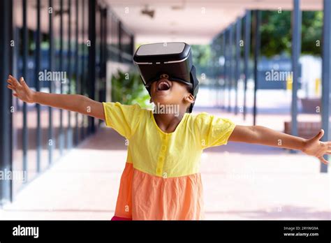 Happy Biracial Schoolgirl Using Vr Headset Standing In Sunny School Corridor Education