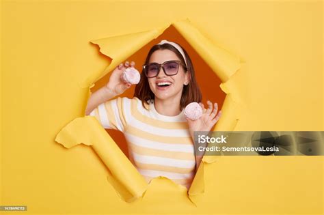 머리띠와 선글라스를 끼고 노란 종이 구멍에 서고 마시멜로를 들고 행복하고 맛있는 단 과자를 먹는 쾌활한 젊은 여성의 실내 사진