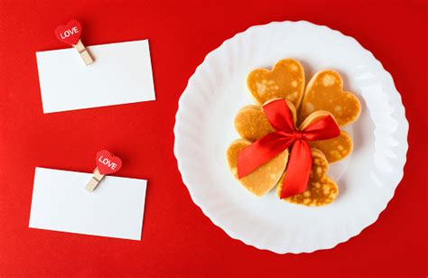 발렌타인 데이를위한 창조적 인 아침 식사 흰색 접시에 하트 모양의 팬케이크 빨간색 새틴 활 빨간색 배경에 텍스트 메모장의 흰색 시트 옆에 복사 공간 0명에 대한 스톡 사진