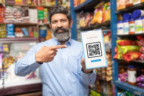 Portrait Of Happy Mature Indian Man Showing Qr Code Scanner To Pay Online At Grocery Shop Or