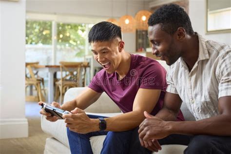 Same Sex Male Couple Or Friends On Sofa At Home With Handheld Mobile Computer Gaming Console